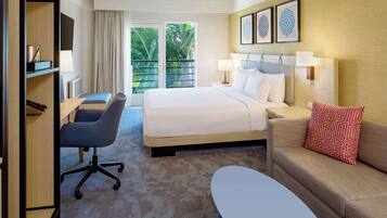Quarto de hotel com cama, escrivaninha, cadeira, sofá e vista para palmeiras pela janela.