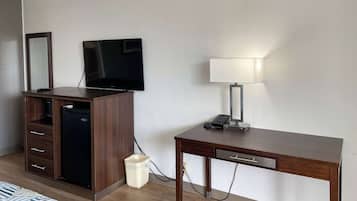 Quarto de hotel com uma escrivaninha de madeira, uma televisão presa a um móvel, uma lixeira e um abajur.