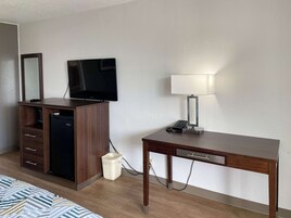 Quarto de hotel com uma escrivaninha de madeira, uma televisão presa a um móvel, uma lixeira e um abajur.