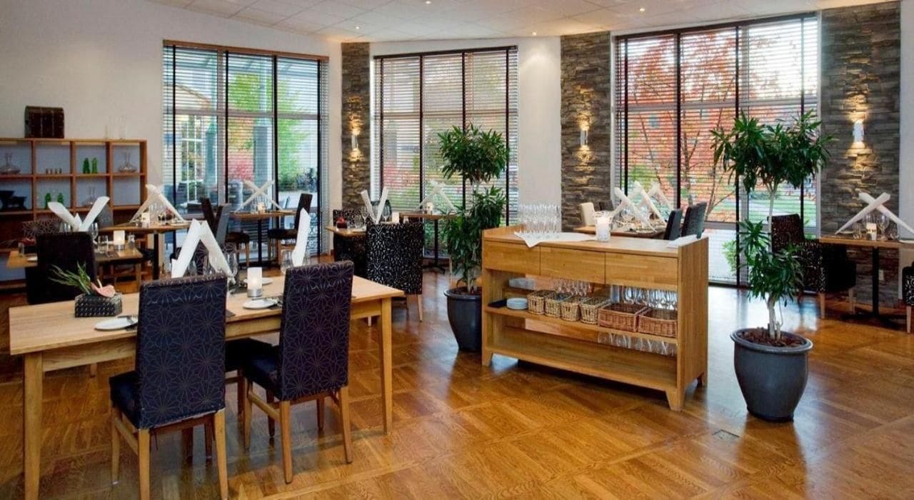 Quarto espaçoso com piso de madeira, uma mesa de jantar central e cadeiras. Há plantas em vasos e uma estante de livros ao fundo.