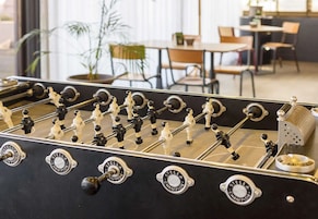 Uma mesa de futebol de mesa com a marcação 'STELLA CHAMPION', em uma sala com cadeiras e uma mesa ao fundo.