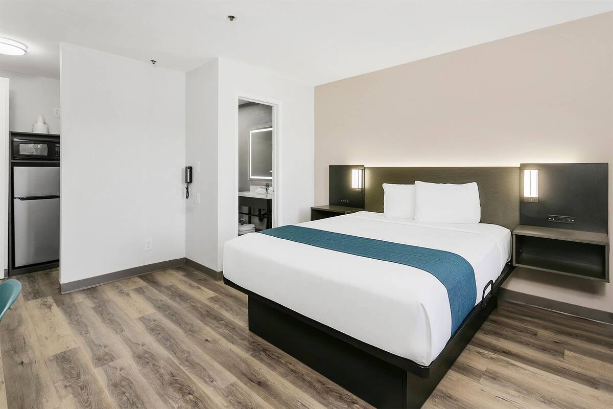 Quarto de hotel moderno com uma cama grande, mesa de cabeceira, televisão integrada e um pequeno espaço de estar.