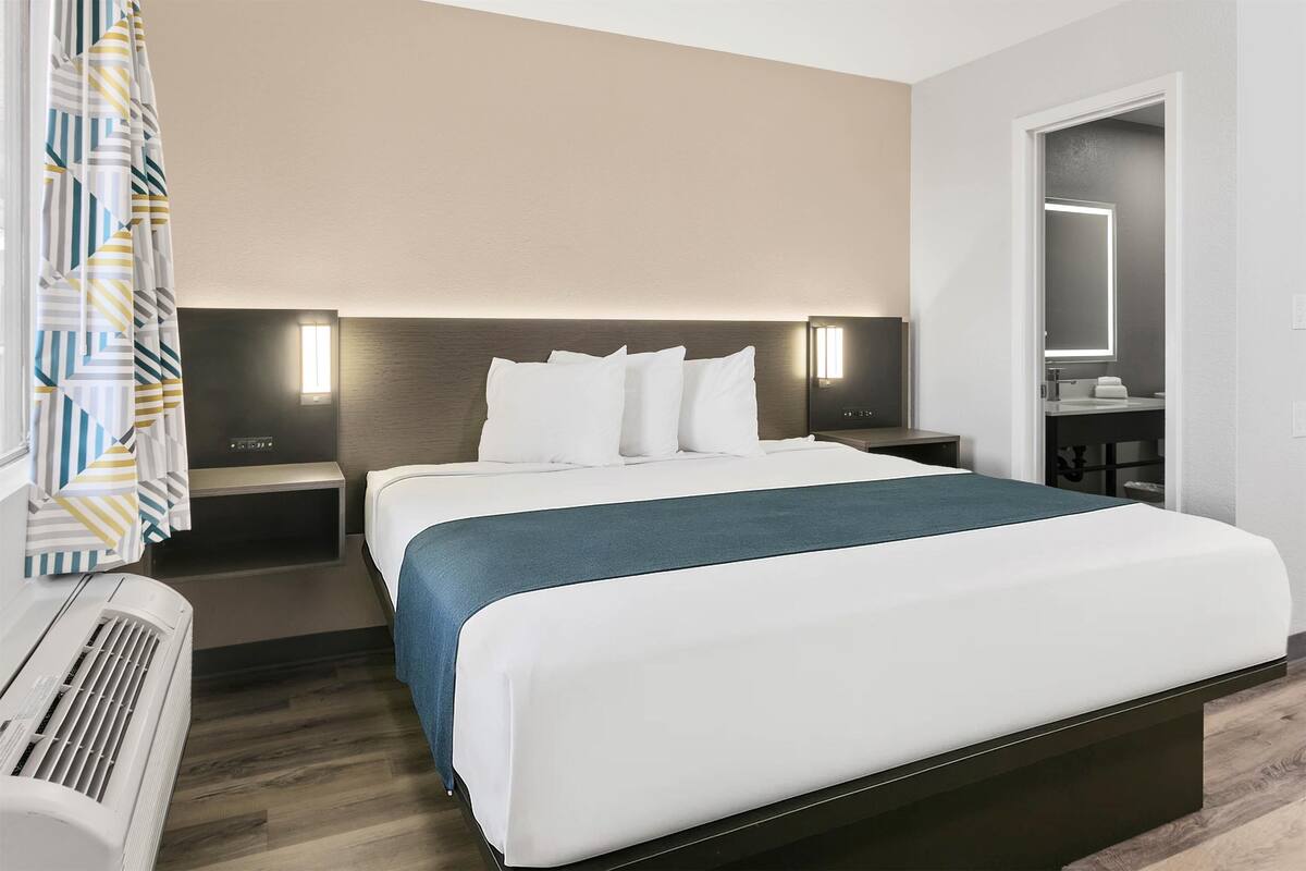 Quarto de hotel com uma cama grande, duas mesas de cabeceira, um criado-mudo, um espelho e um ar condicionado fixado na parede.
