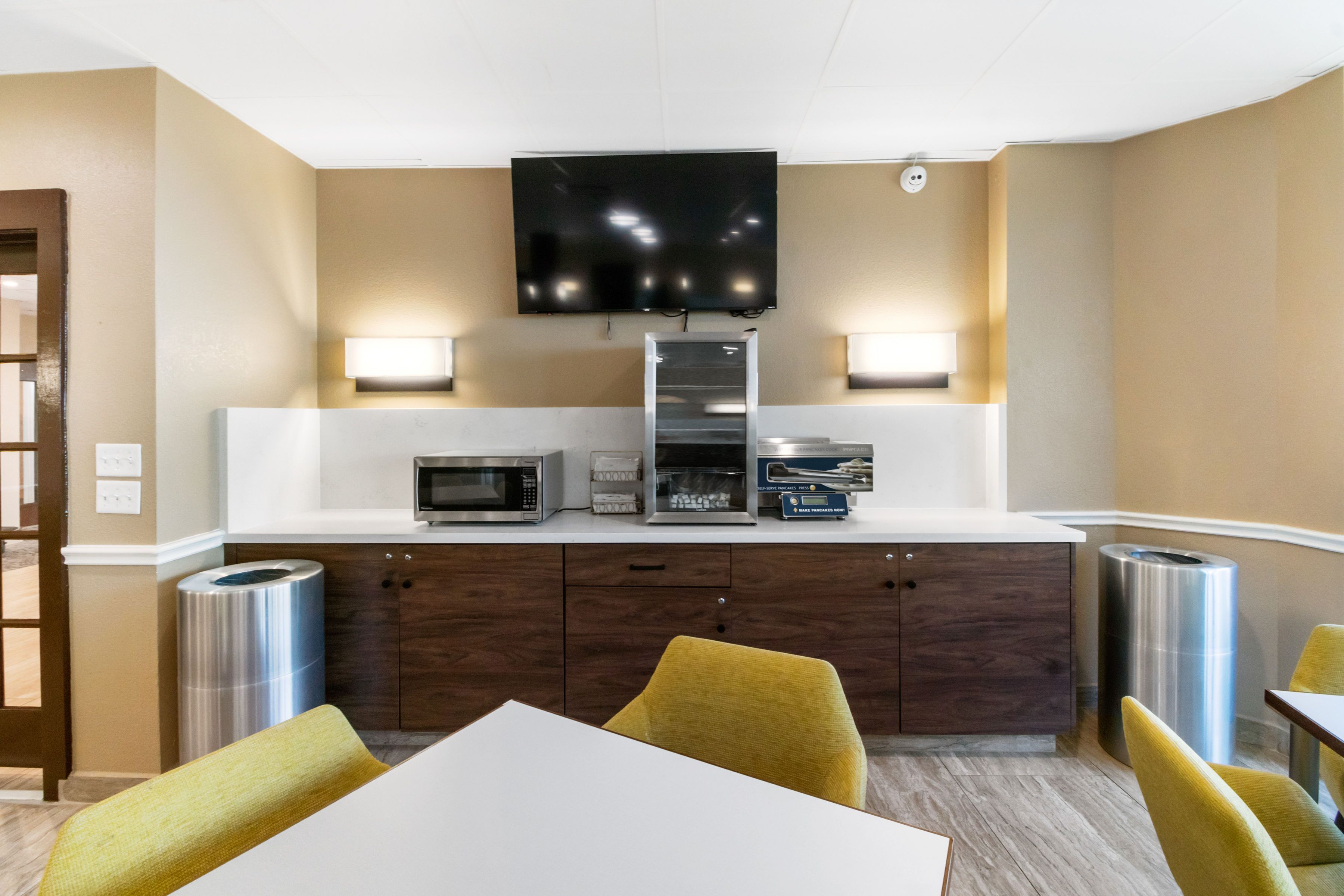 Quarto de hotel com cozinha compacta, televisão de tela plana na parede e área de refeições com cadeiras amarelas.