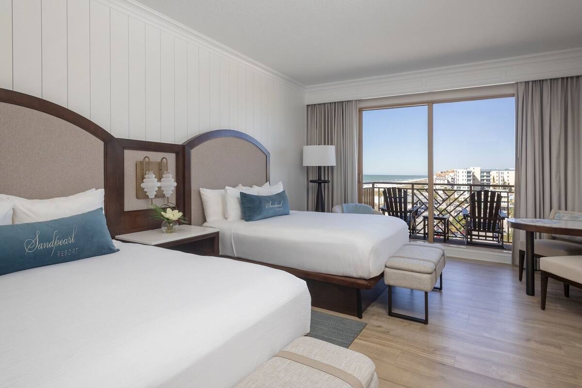 Quarto de hotel com duas camas, uma varanda com vista para a praia e uma escrivaninha com uma cadeira.