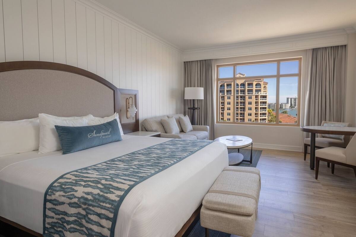 Quarto de hotel com uma cama grande, um sofá, uma mesa pequena e vista para um edifício pela janela.