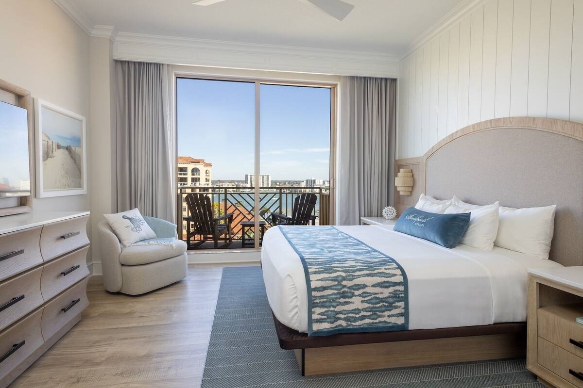 Quarto de hotel com uma cama grande, uma cadeira, uma mesa e vista da cidade pela varanda.