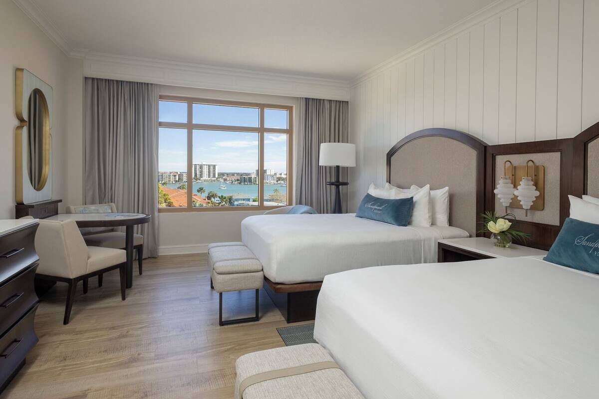 Quarto de hotel com duas camas, uma escrivaninha e uma cadeira. Há uma janela grande com vista para a cidade.