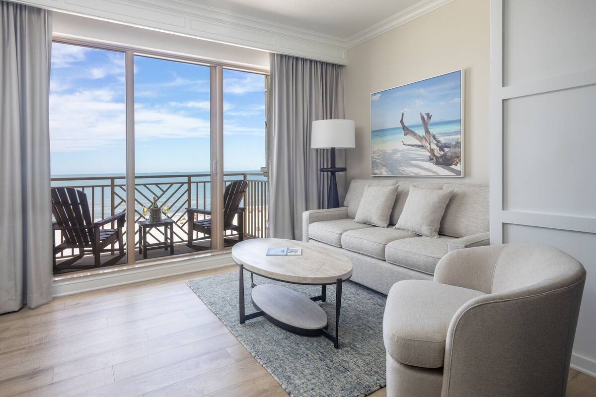 Quarto de hotel com varanda, sofá, mesa de centro e pintura de uma paisagem praiana.