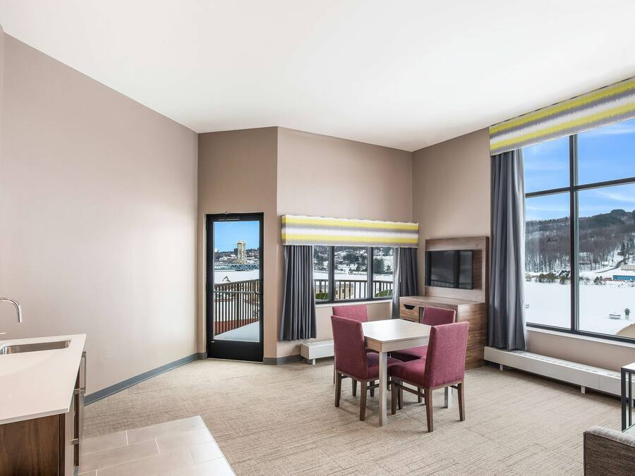 Quarto de hotel com uma janela grande, área de refeições com mesa e cadeiras, televisão e pia.