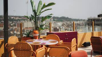Terraço no telhado com cadeiras de vime e uma mesa posta para refeições, com vista para a paisagem urbana.