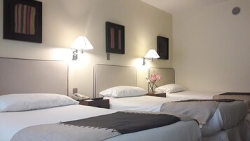 Quarto de hotel com três camas, cada uma com roupa de cama branca e um cobertor escuro. Há abajures e um vaso com flores em uma das camas.