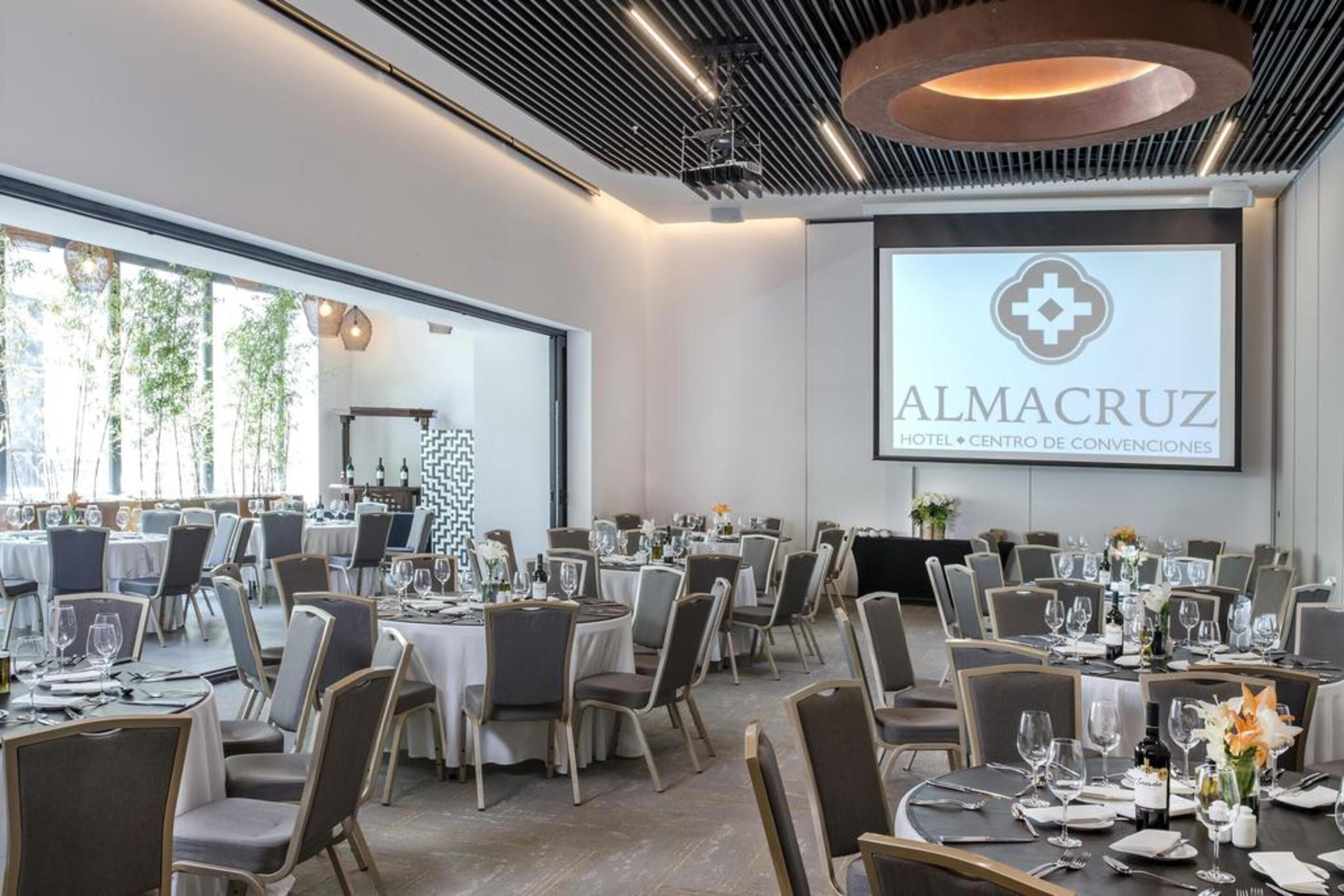 Uma sala de conferências no Hotel Almacruz, com mesas organizadas para uma reunião, uma tela de projetor exibindo o nome do hotel e uma grande janela com luz natural.