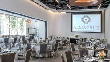 Uma sala de conferências no Hotel Almacruz, com mesas organizadas para uma reunião, uma tela de projetor exibindo o nome do hotel e uma grande janela com luz natural.