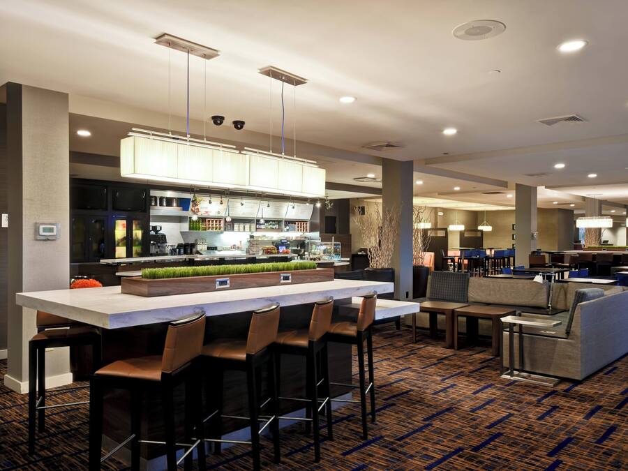 Um lobby de hotel moderno com área de bar central, arranjos de assento e cozinha ao fundo.