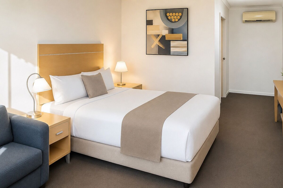Quarto de hotel com cama, mesas de cabeceira, luminária, cadeira e uma obra de arte na parede.