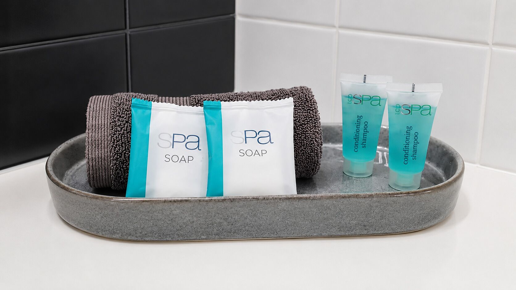 Uma área de pia de banheiro com um descanso cinza de cerâmica, contendo uma toalha enrolada, dois sabonetes e dois frascos de shampoo.