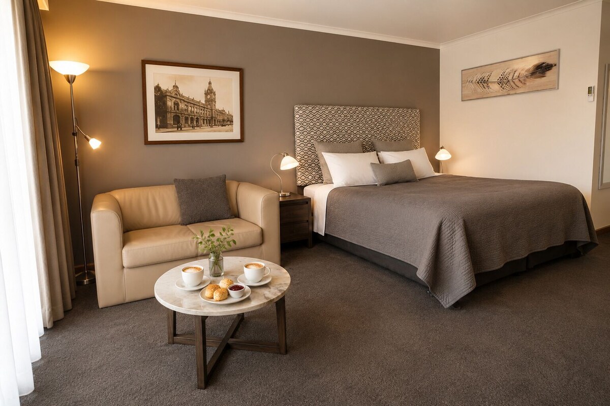 Quarto de hotel com cama, sofá, mesa pequena com um vaso e doces, e uma luminária.