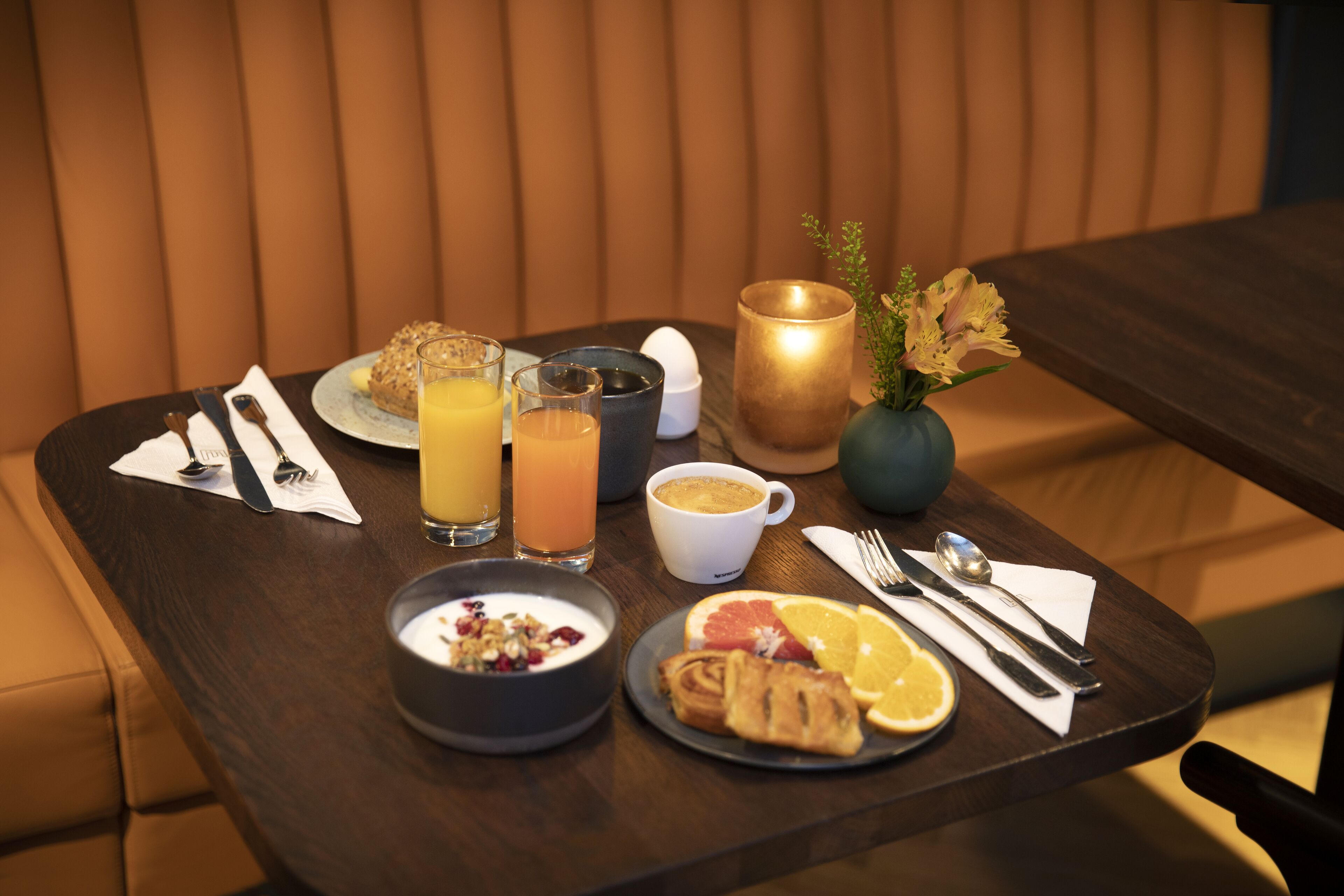 Uma bandeja de café da manhã com um pote de iogurte, um prato com torradas e frutas, uma xícara de café e um copo de suco de laranja sobre uma mesa de madeira em um quarto de hotel.