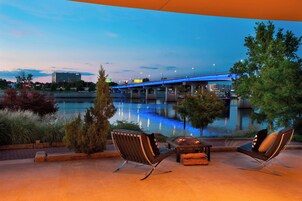 Uma área de estar ao ar livre com duas cadeiras e uma mesa, com vista para um rio e uma ponte ao fundo.