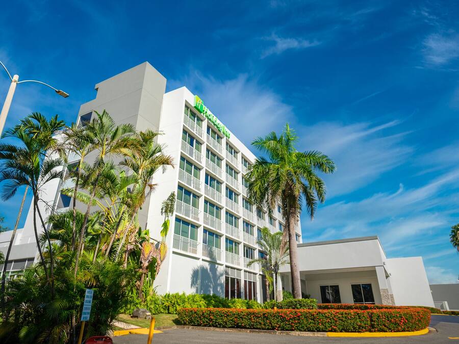 Um hotel moderno com fachada branca, várias varandas e vegetação exuberante, incluindo palmeiras.