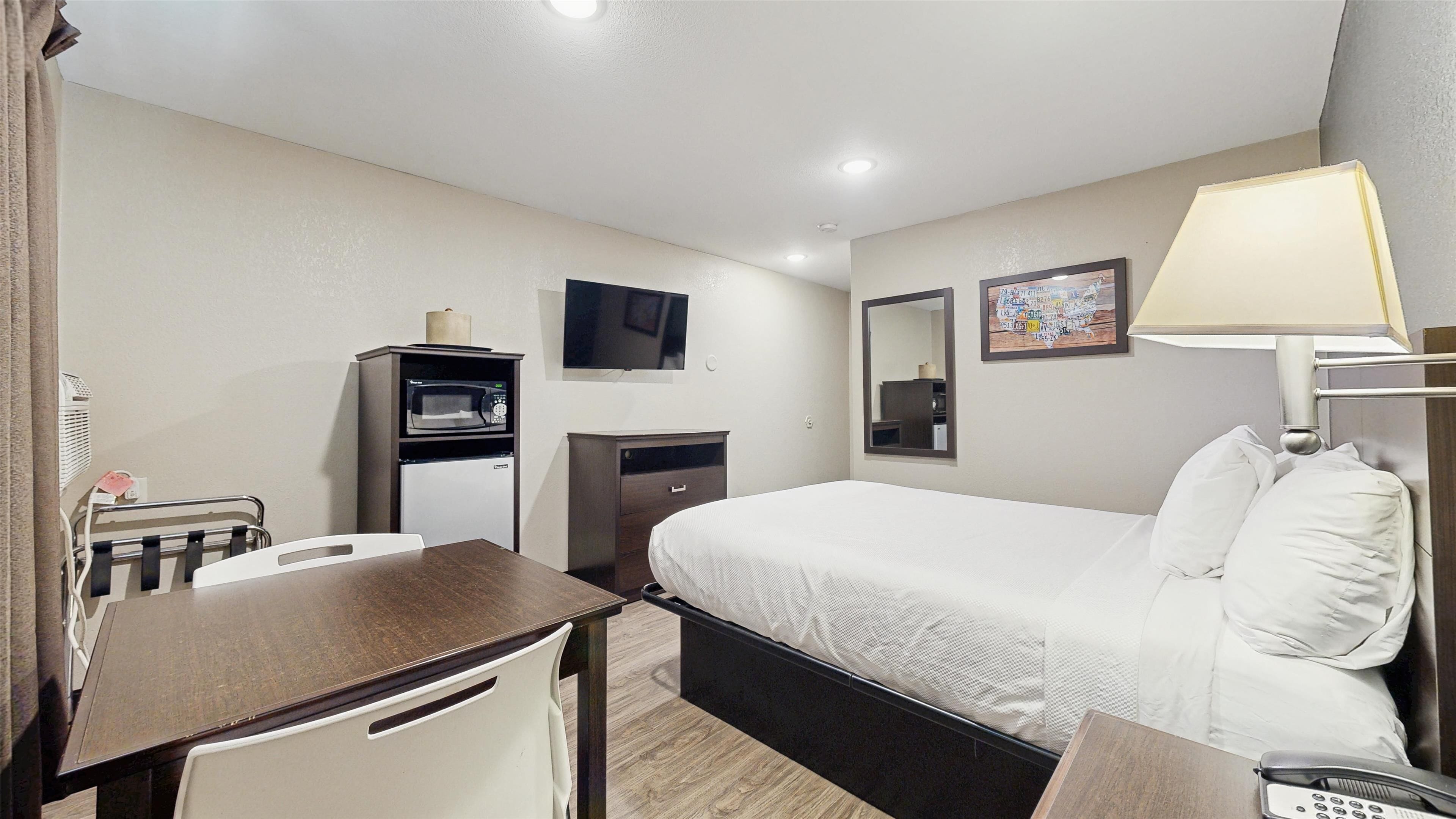 Quarto de hotel com uma cama, escrivaninha, cadeira, micro-ondas, televisão, espelho e telefone.