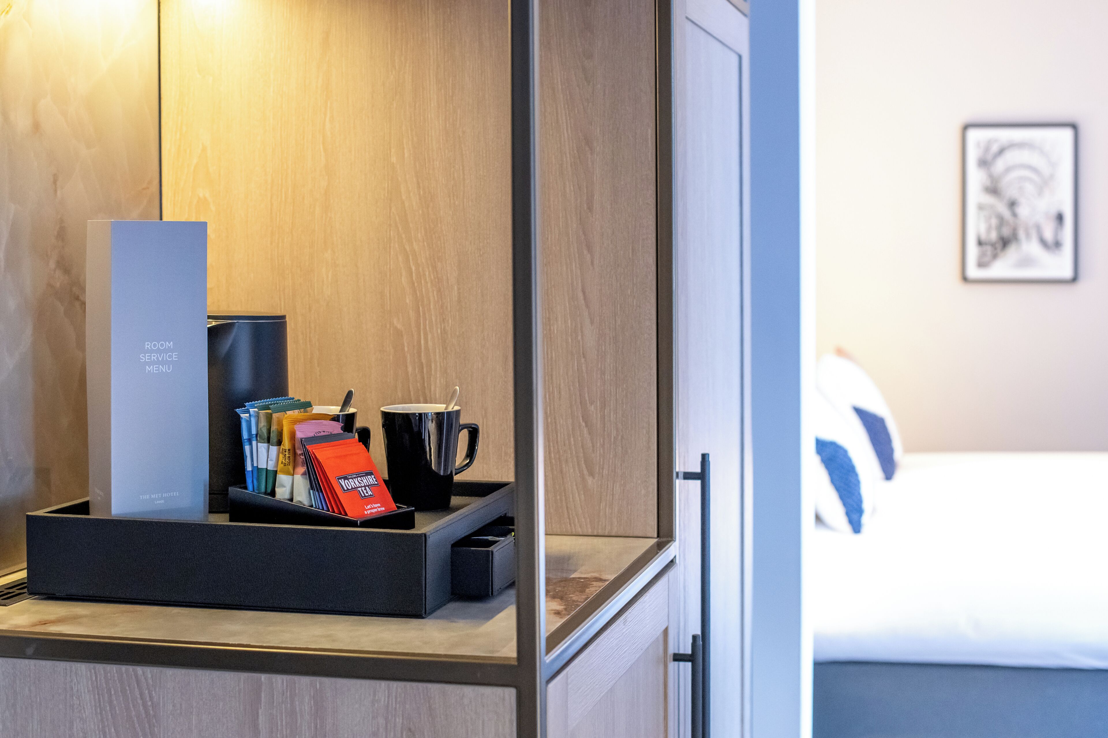 Quarto de hotel com uma mesinha de cabeceira contendo uma cafeteira preta, uma caneca preta, uma embalagem de chá Yorkshire Red e um tabuleiro preto com vários artigos de higiene pessoal.