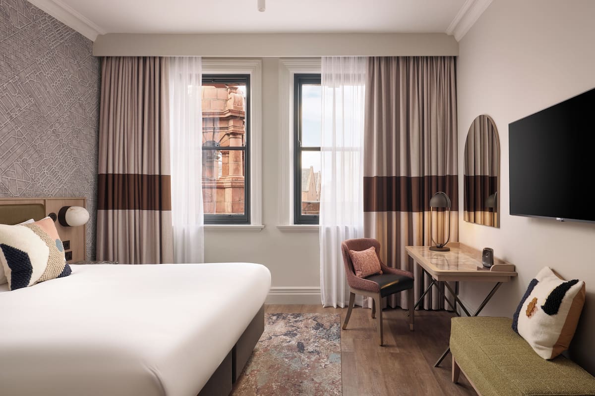 Quarto de hotel com uma cama grande, uma escrivaninha com cadeira, televisão de tela plana e vista para a cidade pelas janelas.
