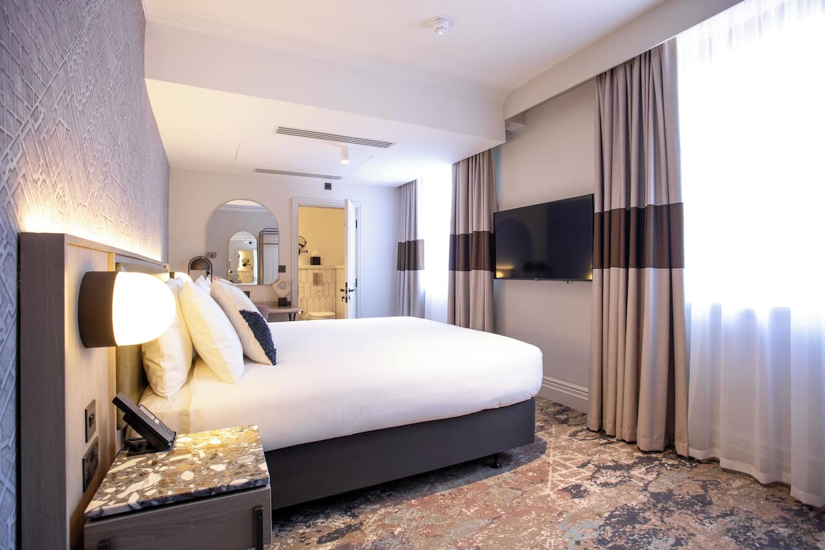 Quarto de hotel moderno com uma cama grande, uma mesa de cabeceira com luminária, uma TV de tela plana na parede e uma grande janela com cortinas.