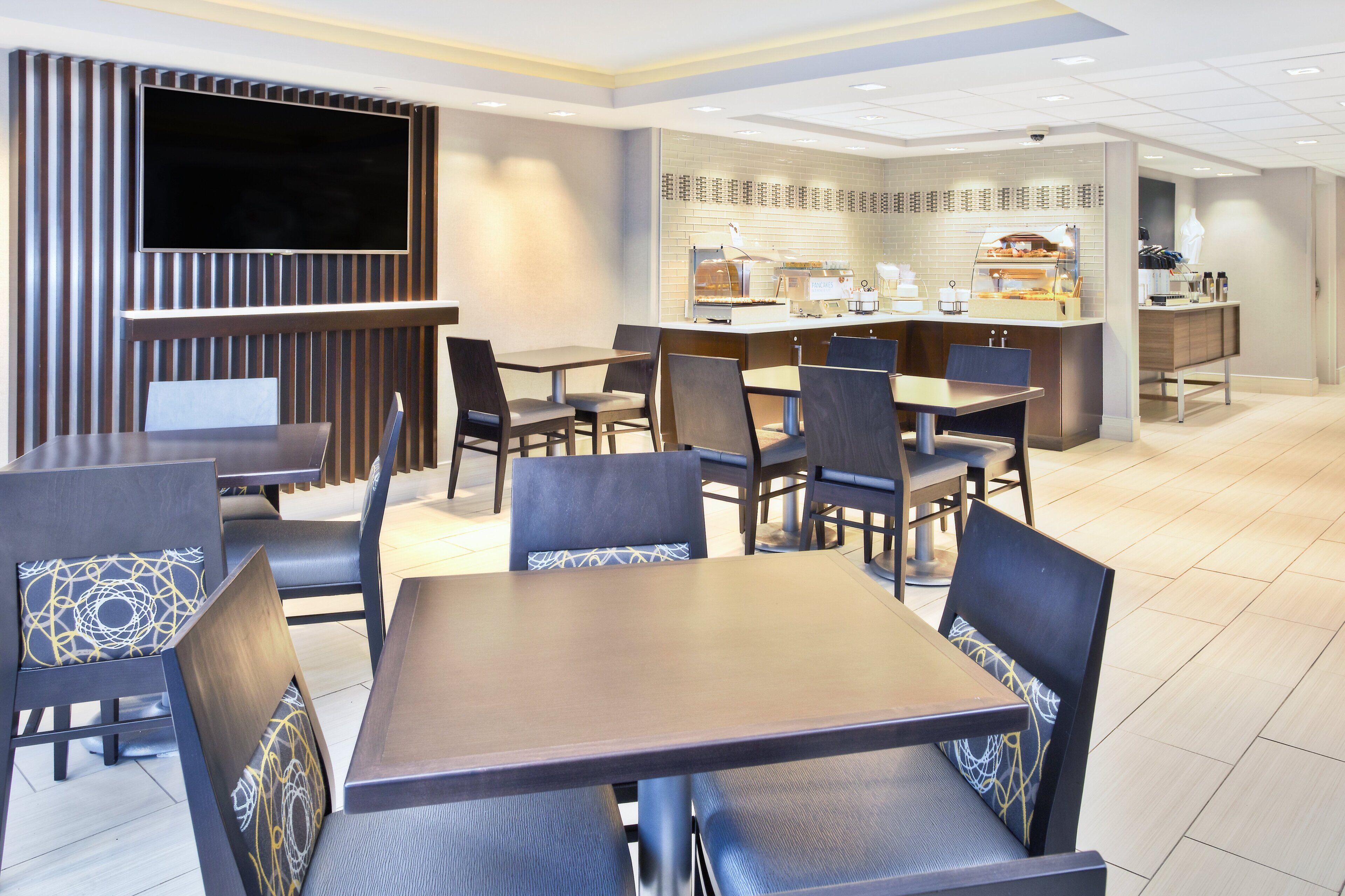 Uma sala de jantar de hotel com mesas, cadeiras, uma televisão na parede e um balcão de café da manhã com diversos alimentos.