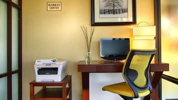 Um espaço de trabalho com impressora, monitor de computador e uma cadeira com luminária. Um quadro e uma placa com a inscrição "BUSINESS CENTER" estão na parede.