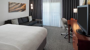 Quarto de hotel com uma cama grande, um sofá, uma escrivaninha com televisão e uma janela com cortinas.