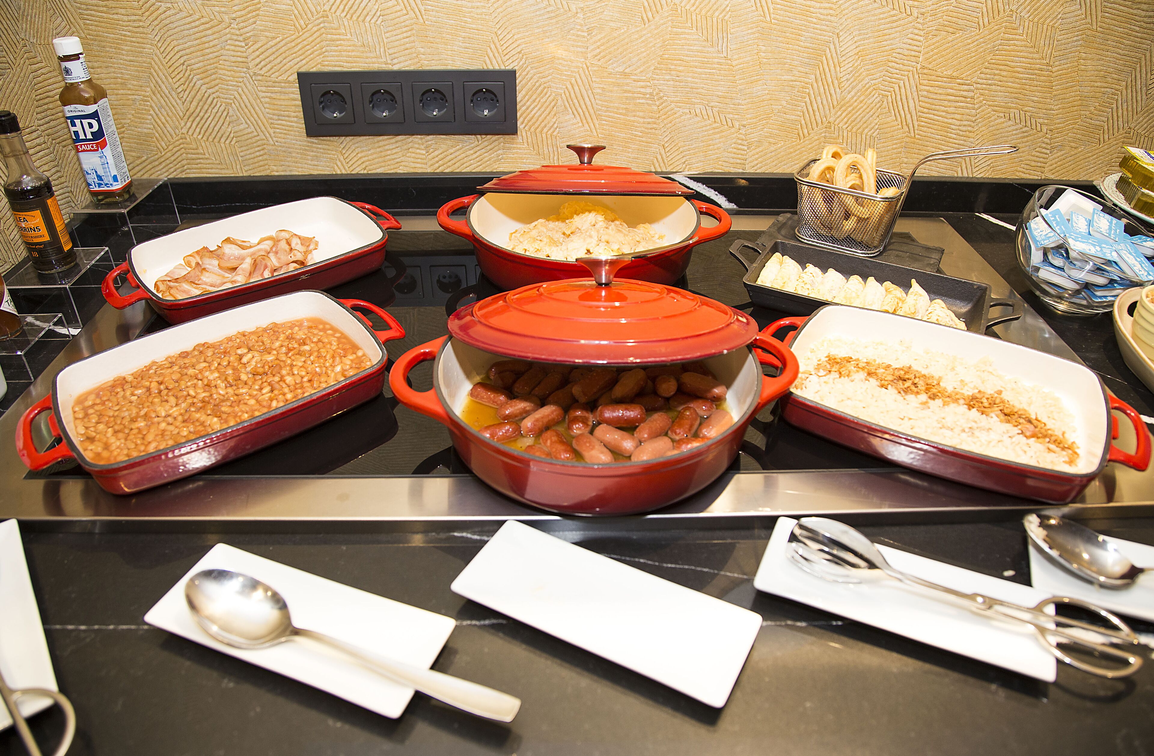 Uma mesa de buffet com diversos pratos em travessas de cerâmica vermelha e branca, incluindo feijão cozido, linguiça e arroz.
