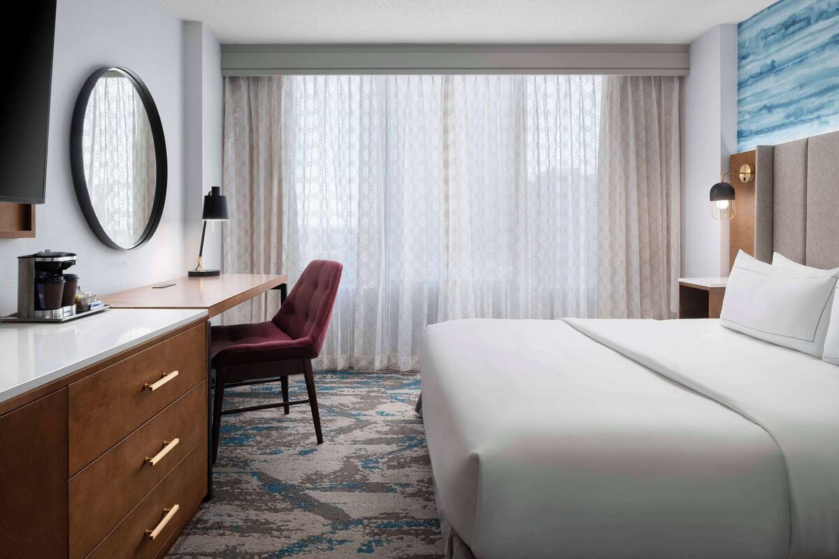 Quarto de hotel com uma cama, uma escrivaninha com cadeira, máquina de café, espelho e janela com cortinas.