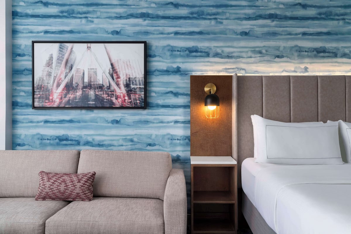 Quarto de hotel com cama, mesa de cabeceira, quadro com vista urbana e sofá com almofada estampada.