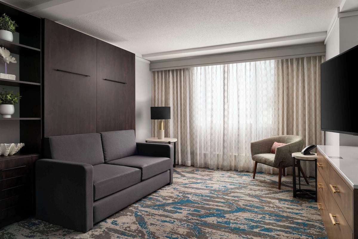 Quarto de hotel moderno com sofá cinza, uma pequena mesa redonda com abajur, televisão, cadeira e uma grande janela com cortinas.