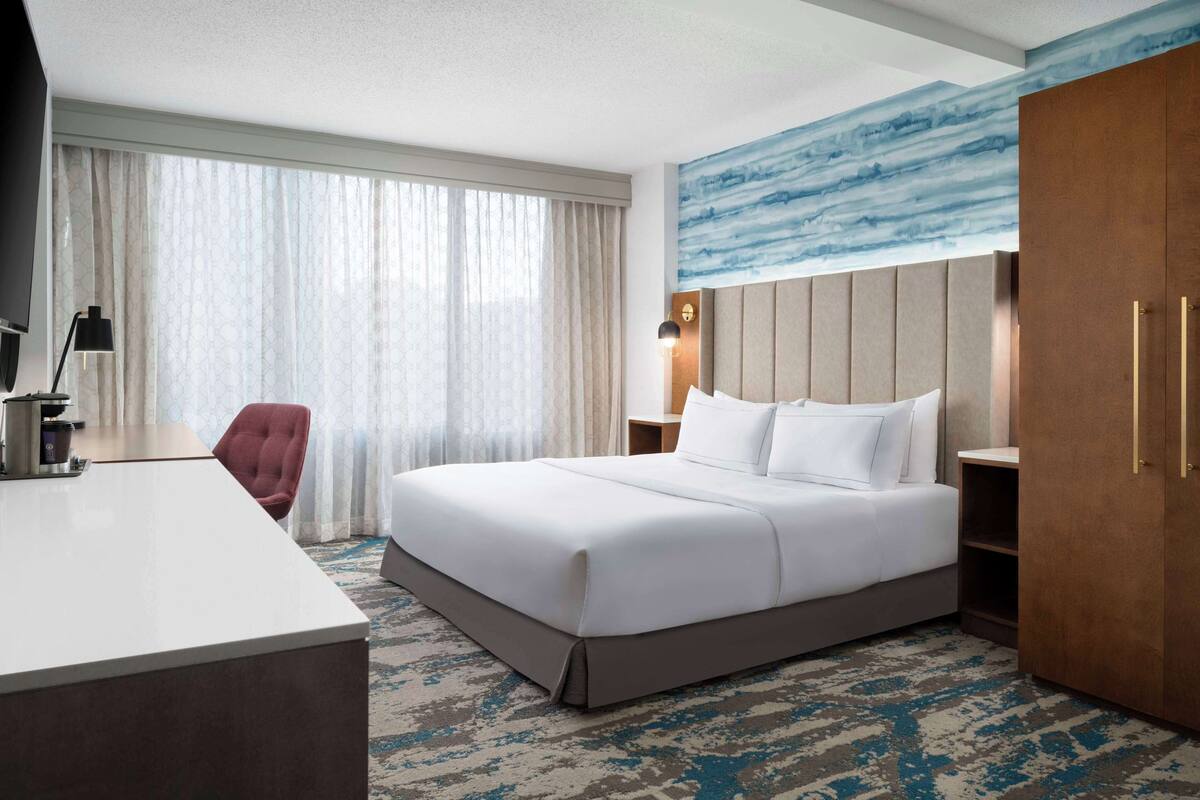 Quarto de hotel com uma cama grande, escrivaninha, cadeira, guarda-roupa e janela com cortinas.