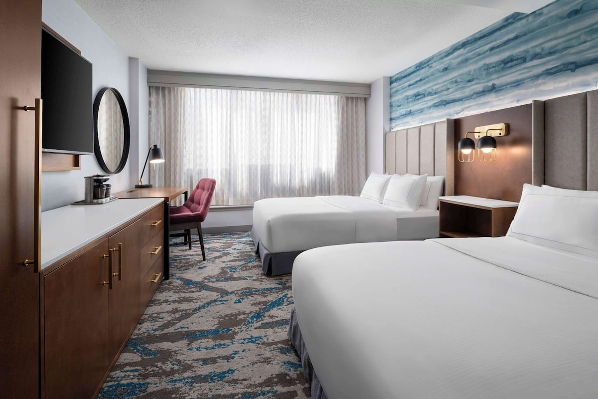 Quarto de hotel com duas camas, uma escrivaninha, uma cadeira, uma televisão e uma janela com cortinas.