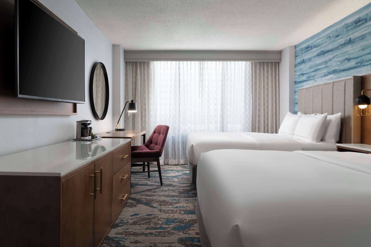 Quarto de hotel com televisão de tela plana, escrivaninha com luminária, uma cadeira, duas camas e uma janela grande com cortinas.