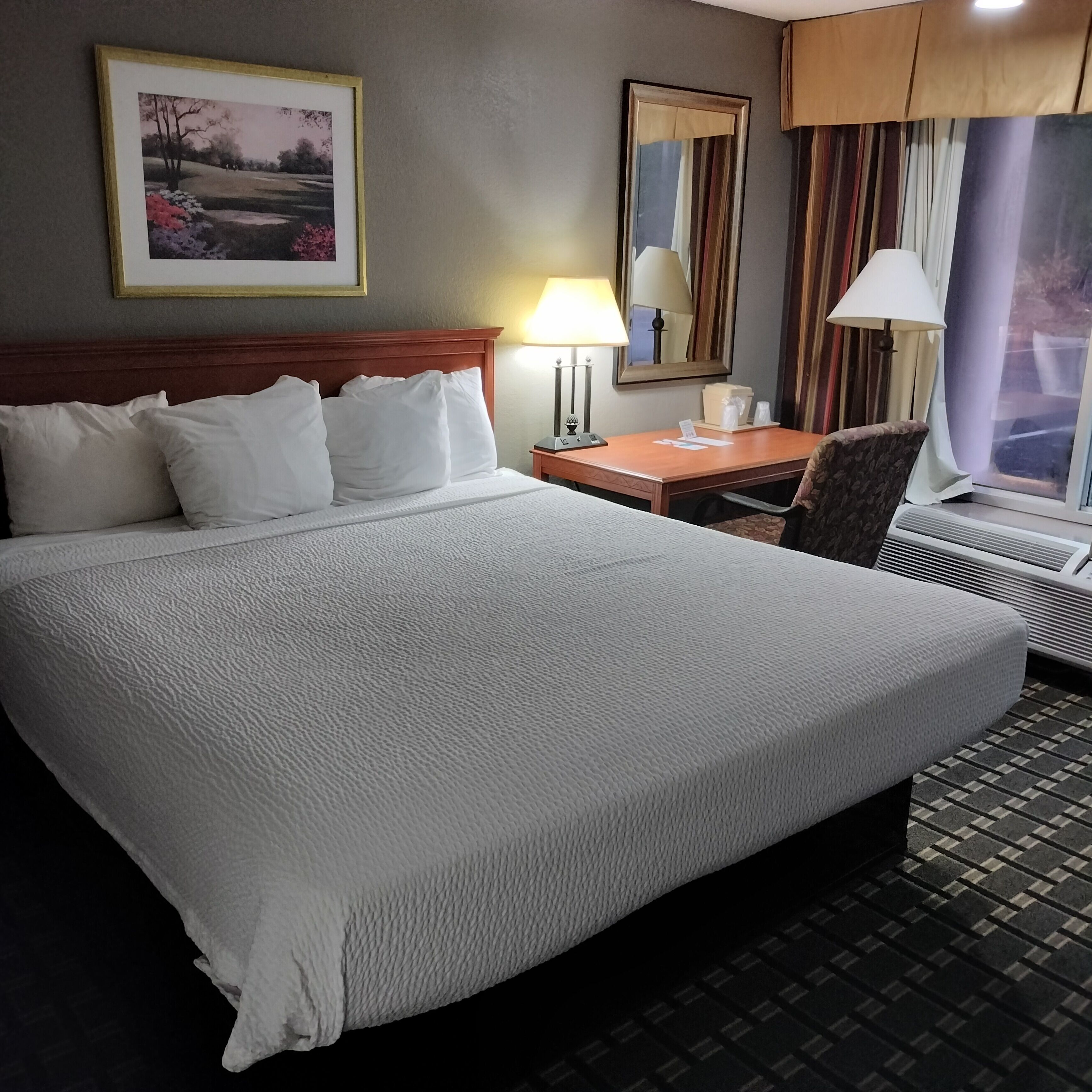 Quarto de hotel com uma cama grande, uma escrivaninha, uma cadeira, uma luminária, um espelho e uma janela com cortinas.