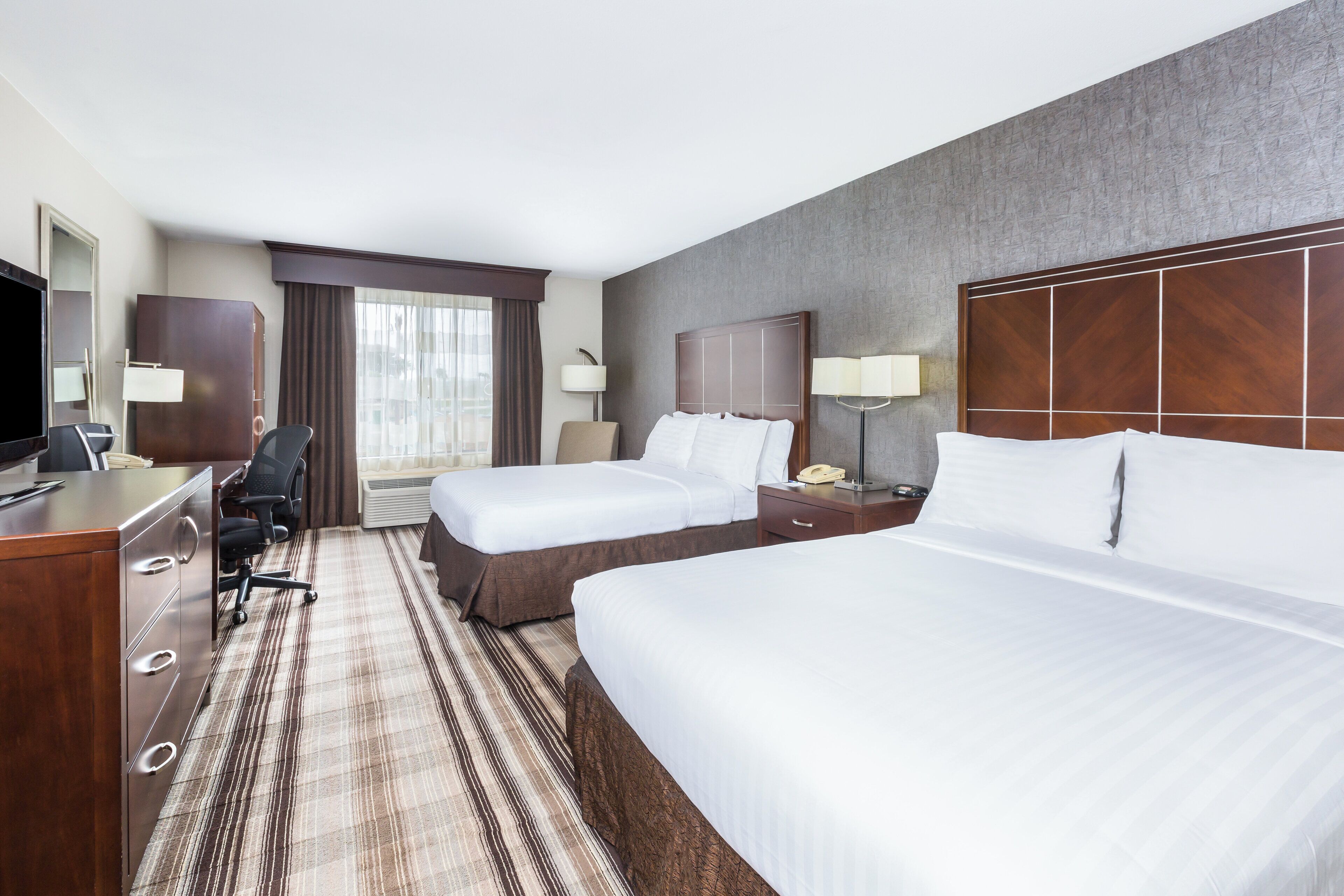 Quarto de hotel com duas camas, uma escrivaninha, uma cadeira, uma televisão e uma janela com cortinas.