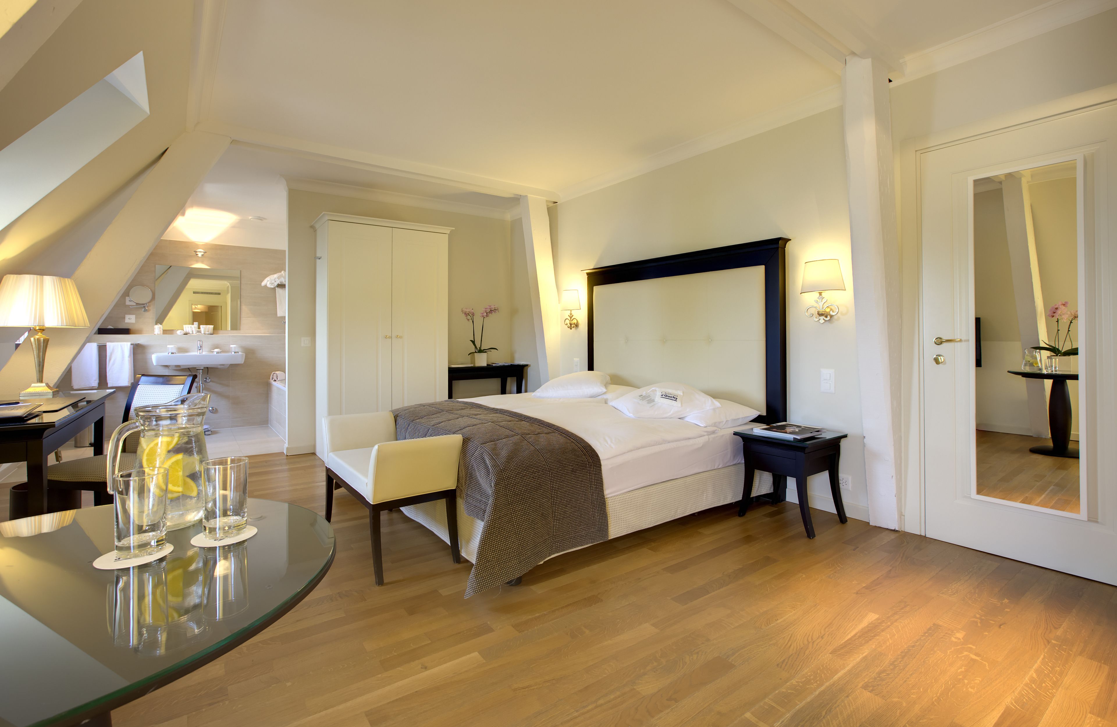Quarto de hotel com cama, mesas de cabeceira, escrivaninha com abajur, uma cadeira e uma mesa de vidro com jarra e copos.