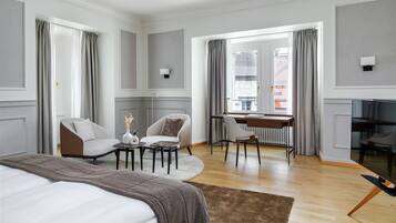 Quarto de hotel moderno com uma cama grande, duas poltronas, uma pequena mesa com um vaso, uma escrivaninha com cadeira, uma TV e vista para um edifício através de amplas janelas.