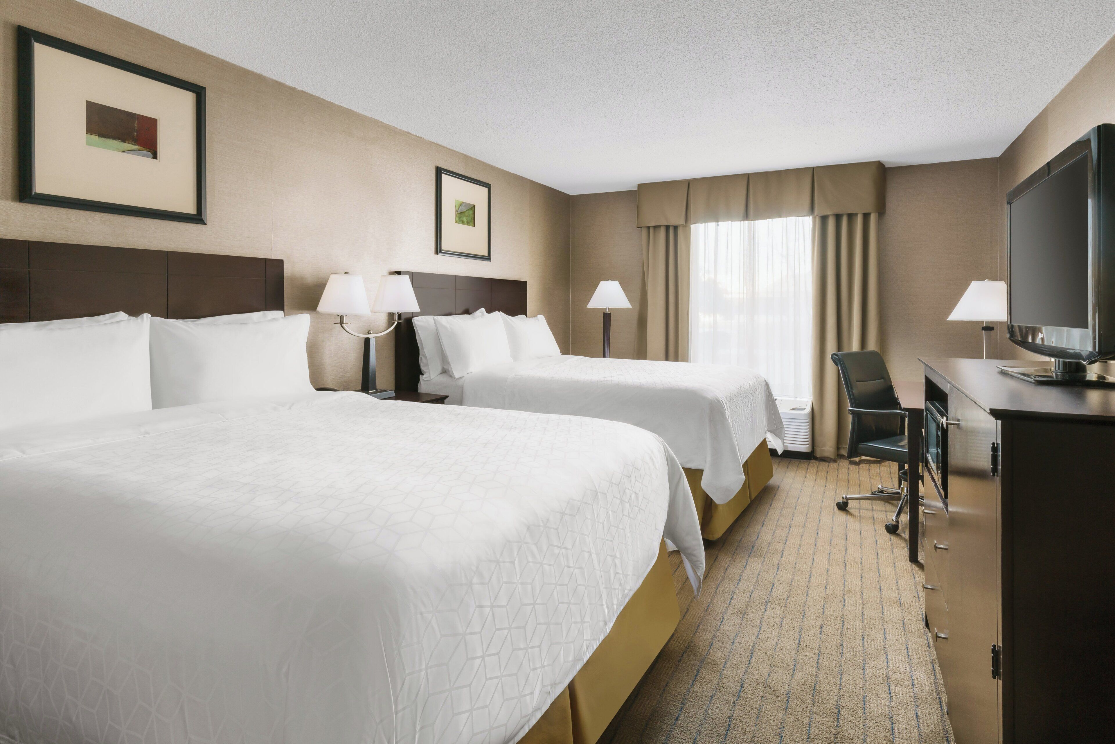 Quarto de hotel com duas camas, uma escrivaninha, uma cadeira, uma televisão e duas luminárias.