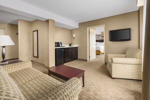 Quarto de hotel com área de estar, cozinha compacta, televisão e cama.