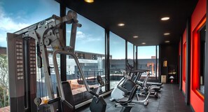 Academia com equipamentos de exercício, janelas amplas e vista para edifícios.