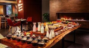 Disposição de buffet com diversos alimentos e bebidas, uma lareira e arranjos de assentos.