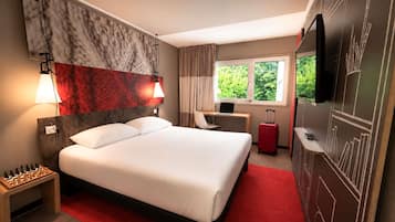 Quarto de hotel com cama, escrivaninha, televisão, janela com vista para áreas verdes e tapete vermelho.