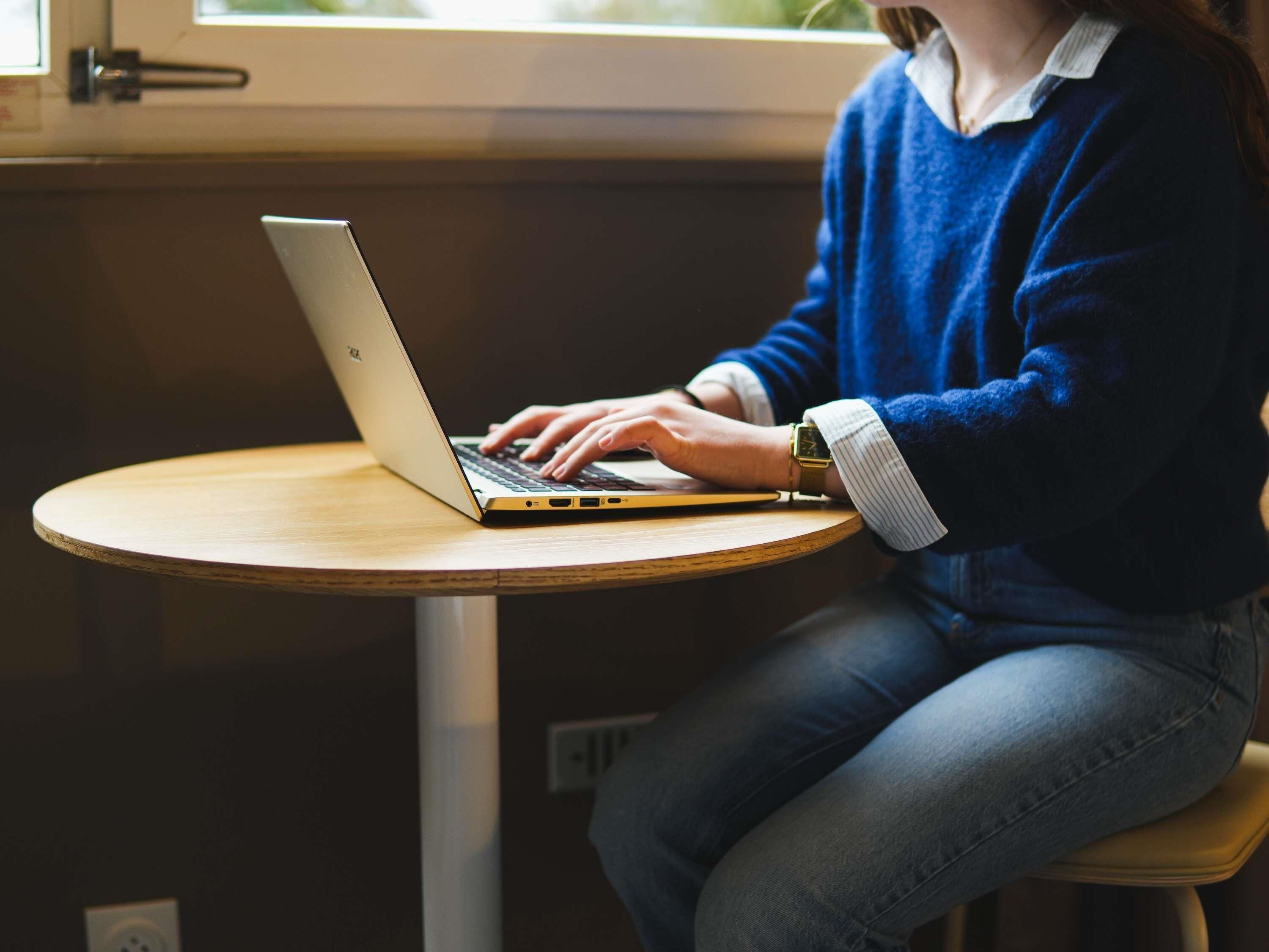 Uma pessoa usando um laptop em cima de uma mesa redonda de madeira num quarto com janela e porta.