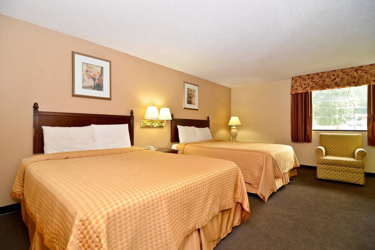 Quarto de hotel com duas camas, uma cadeira e duas luminárias fixadas na parede.